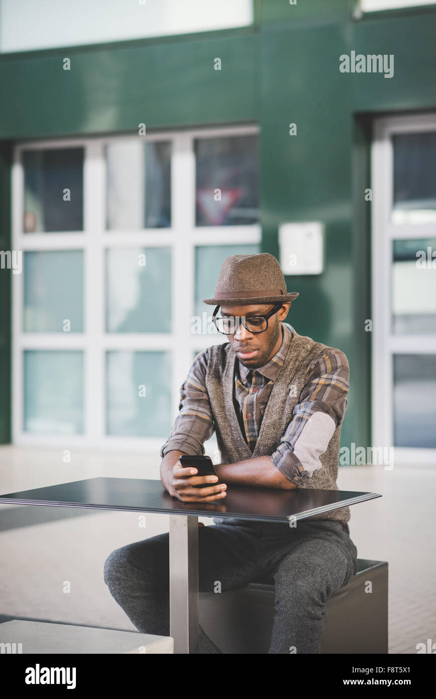 young handsome afro black man sitting on a table, smartphone handhold, looking down the screen ...
