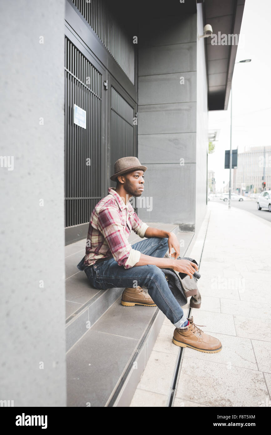 young handsome afro black man sitting on a staircase, smartphone handheld, looking straight ...
