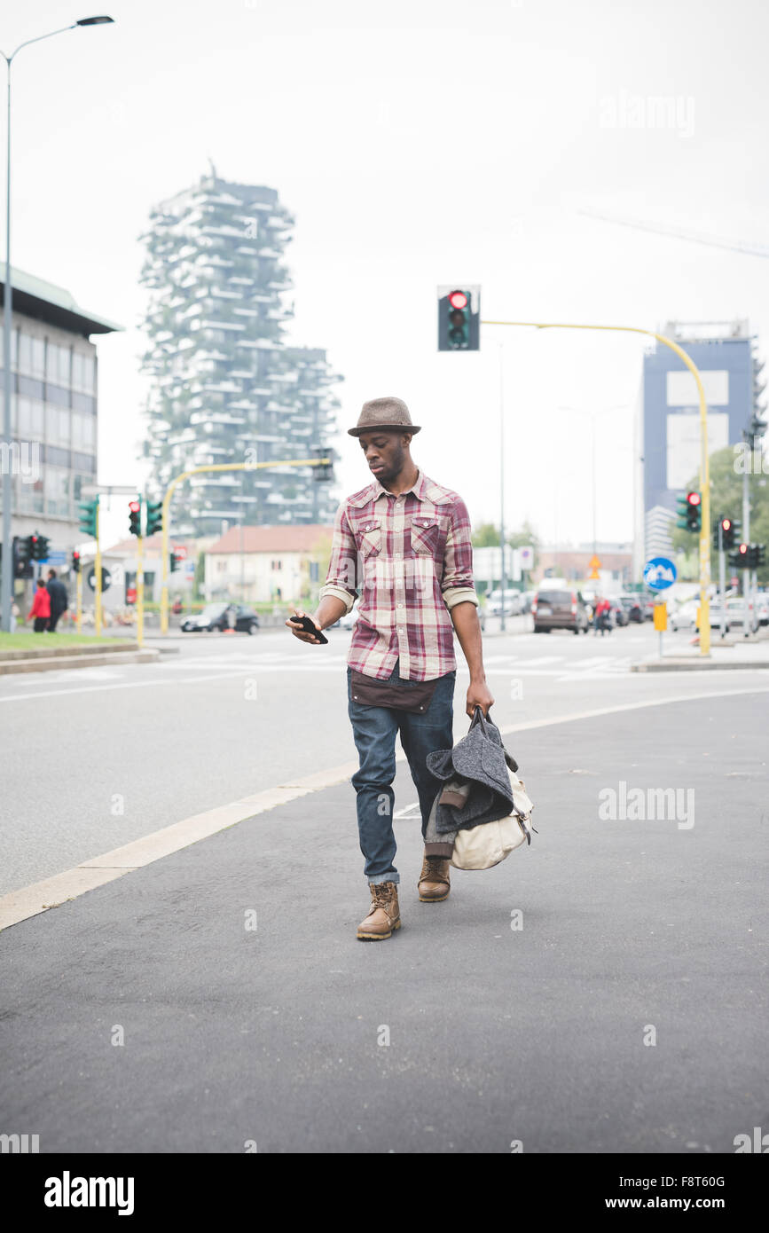 Young handsome afro black man walking in the street of the city, holding a bag, using a ...