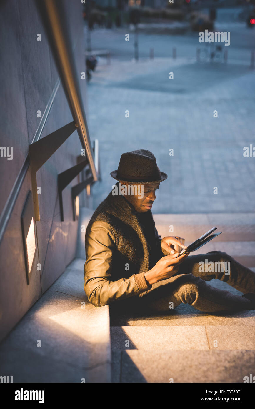 young handsome afro black man sitting on a staircase, using a tablet, looking downward the ...