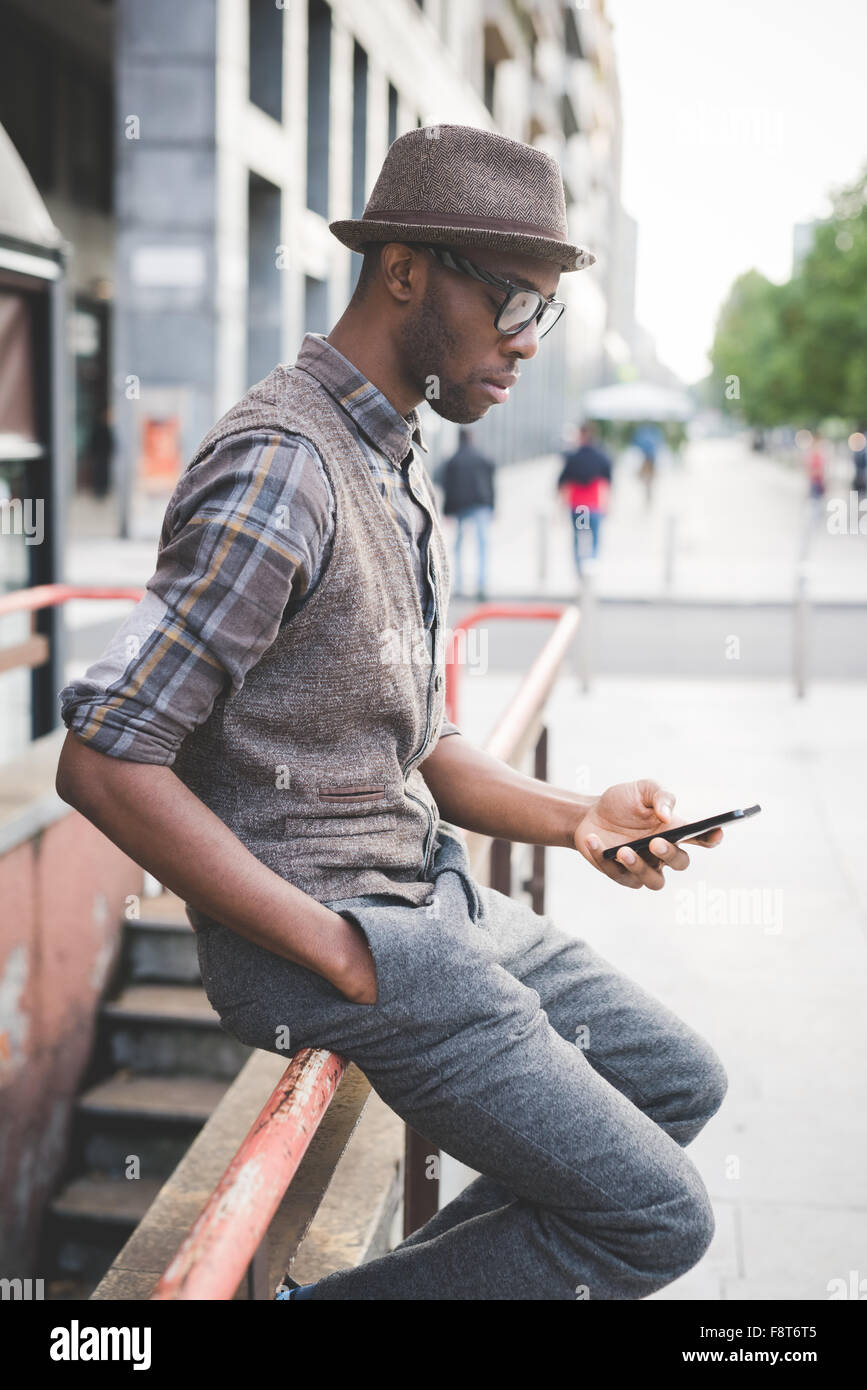 young handsome afro black man sitting on a handrail outside in the city using a smartphone ...