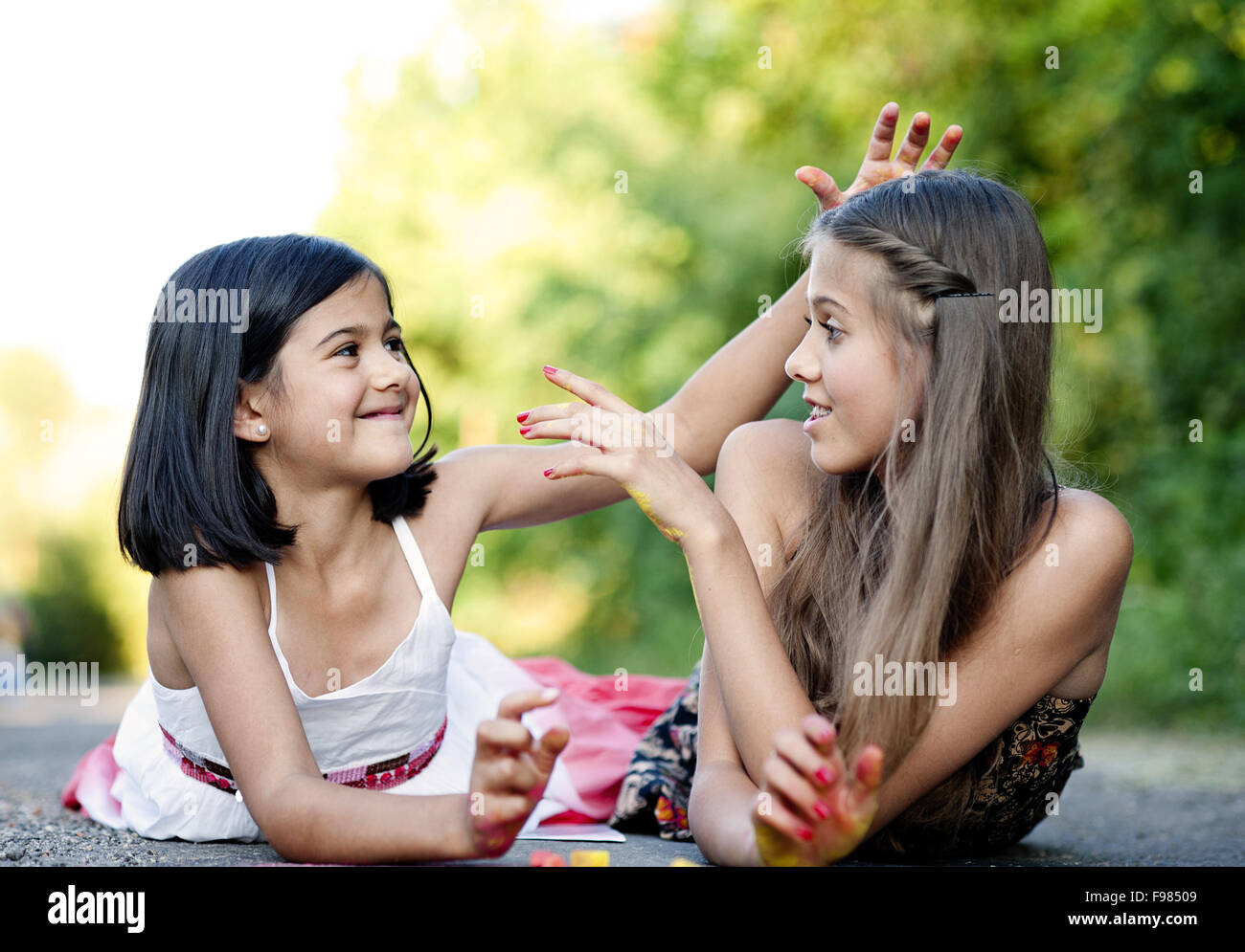 Two sisters laughing and playing with chalks on pavement in green sunny ...