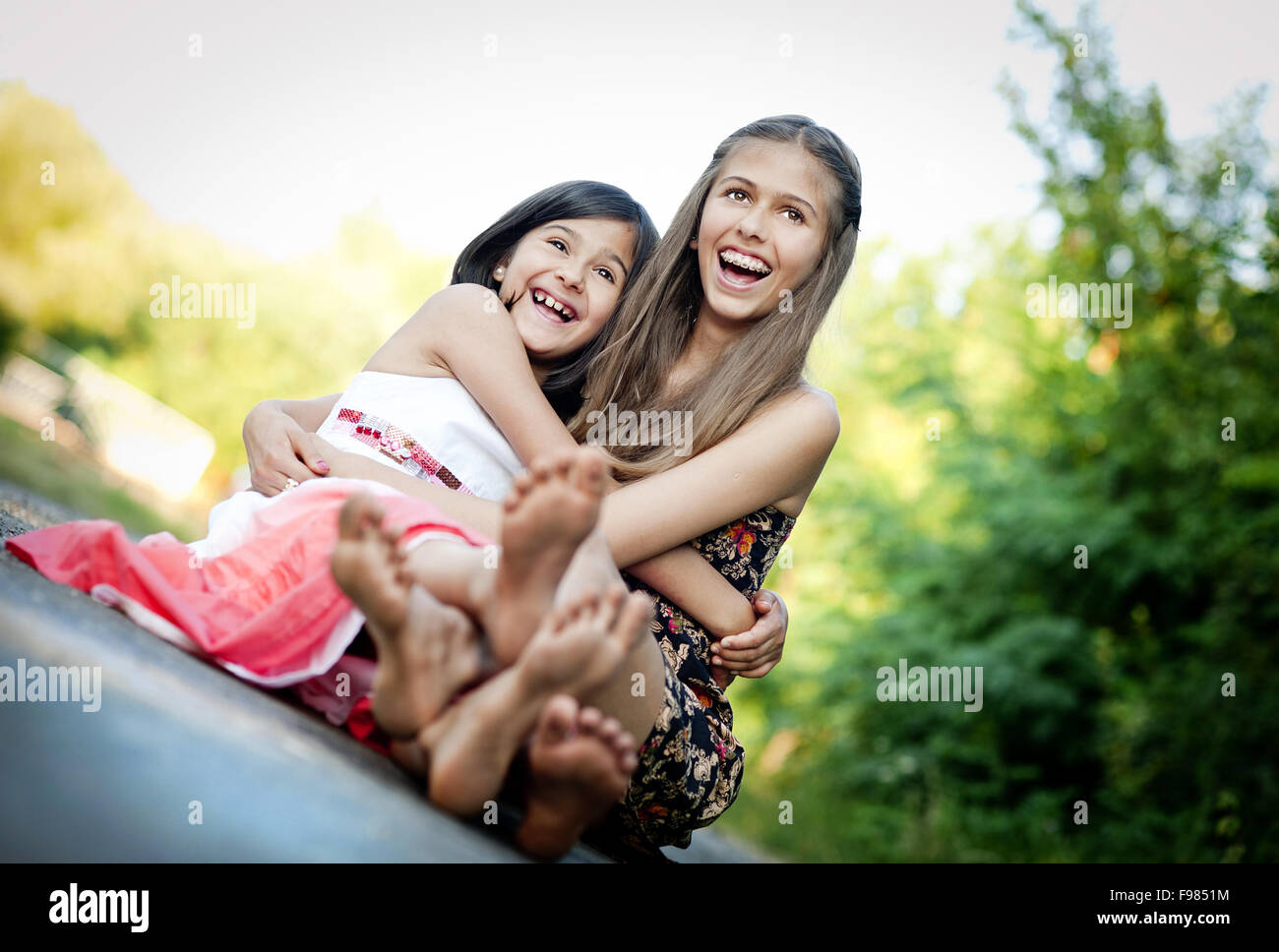 Two sisters laughing and hugging on pavement in green sunny park Stock ...