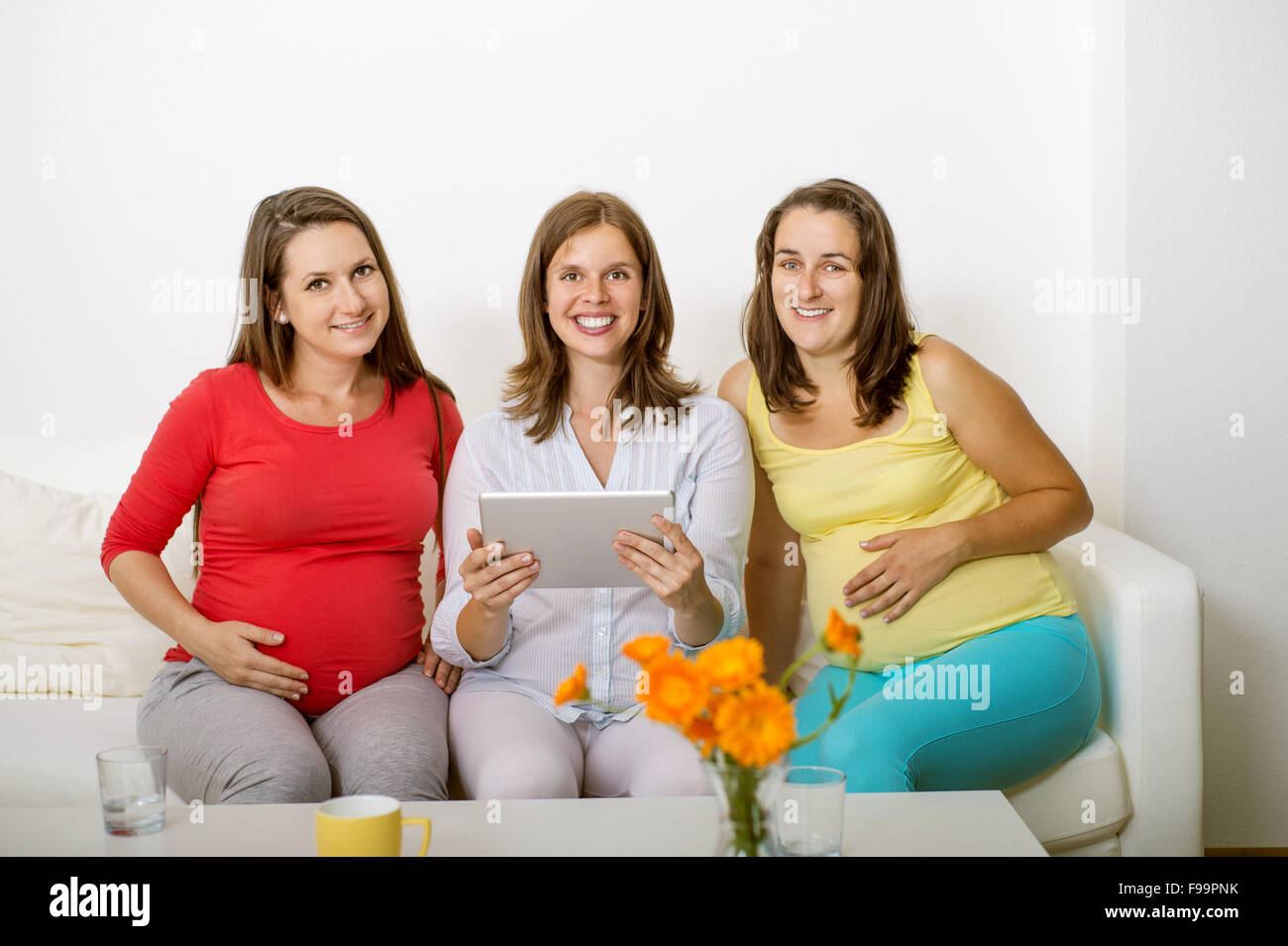 Three pregnant women sitting on sofa, chatting and using digital tablet ...
