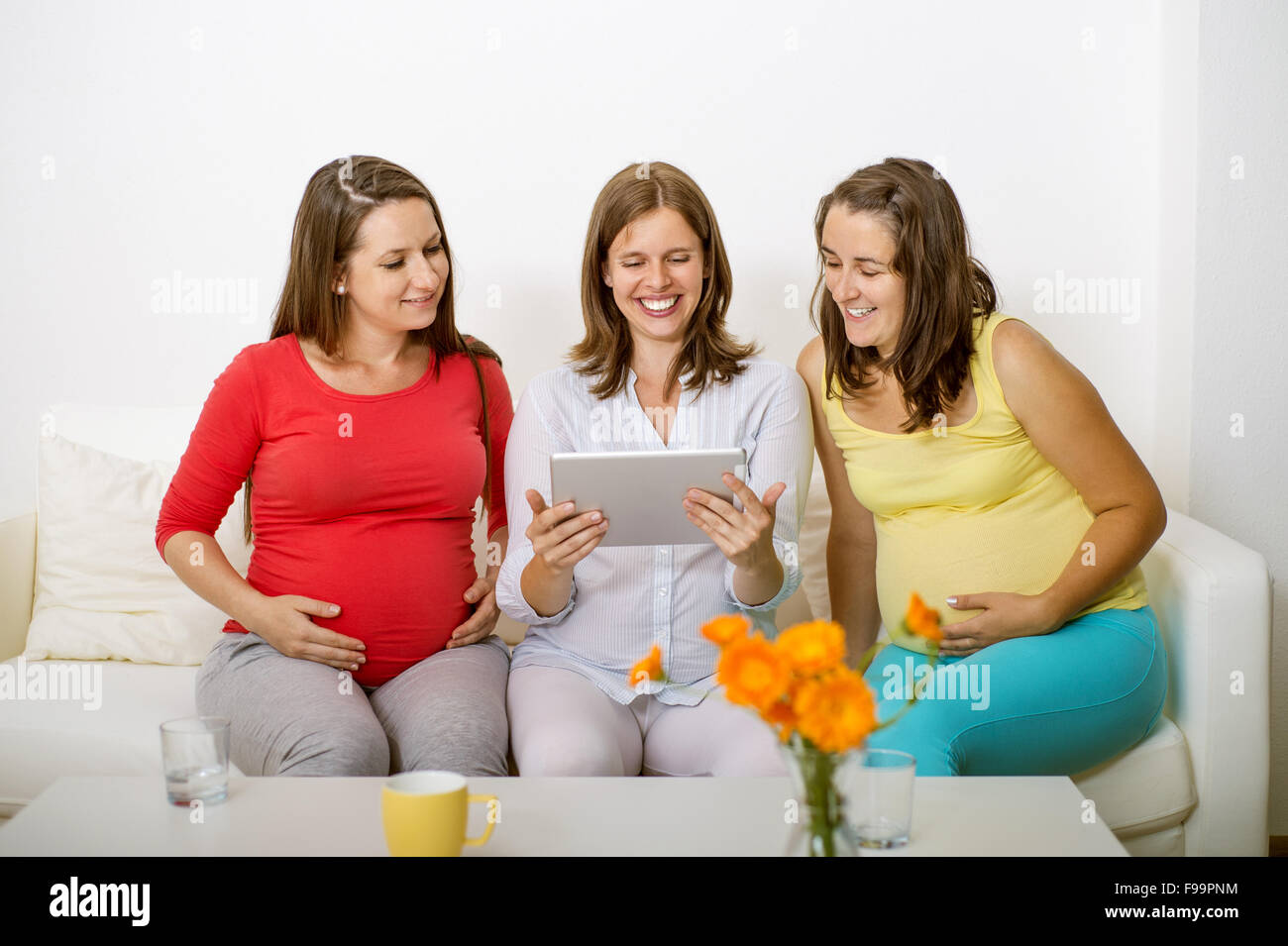 Three pregnant women sitting on sofa, chatting and using digital tablet ...