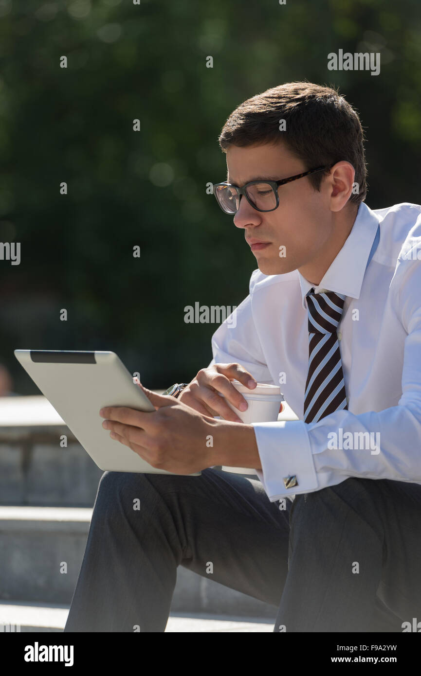 Young man drinking coffee outdoors at park and using tablet computer ...
