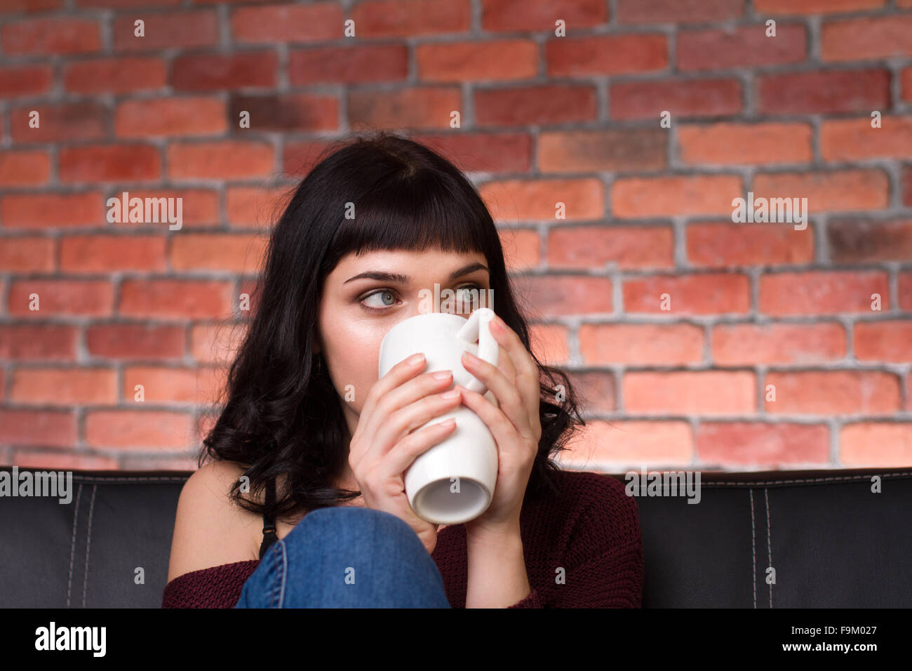 Pretty cute young woman drinking hot tea on sofa over brick wall ...