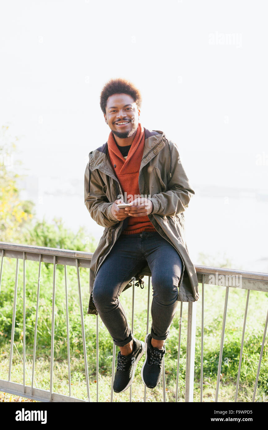 Portrait of smiling young man with smartphone sitting on a railing ...