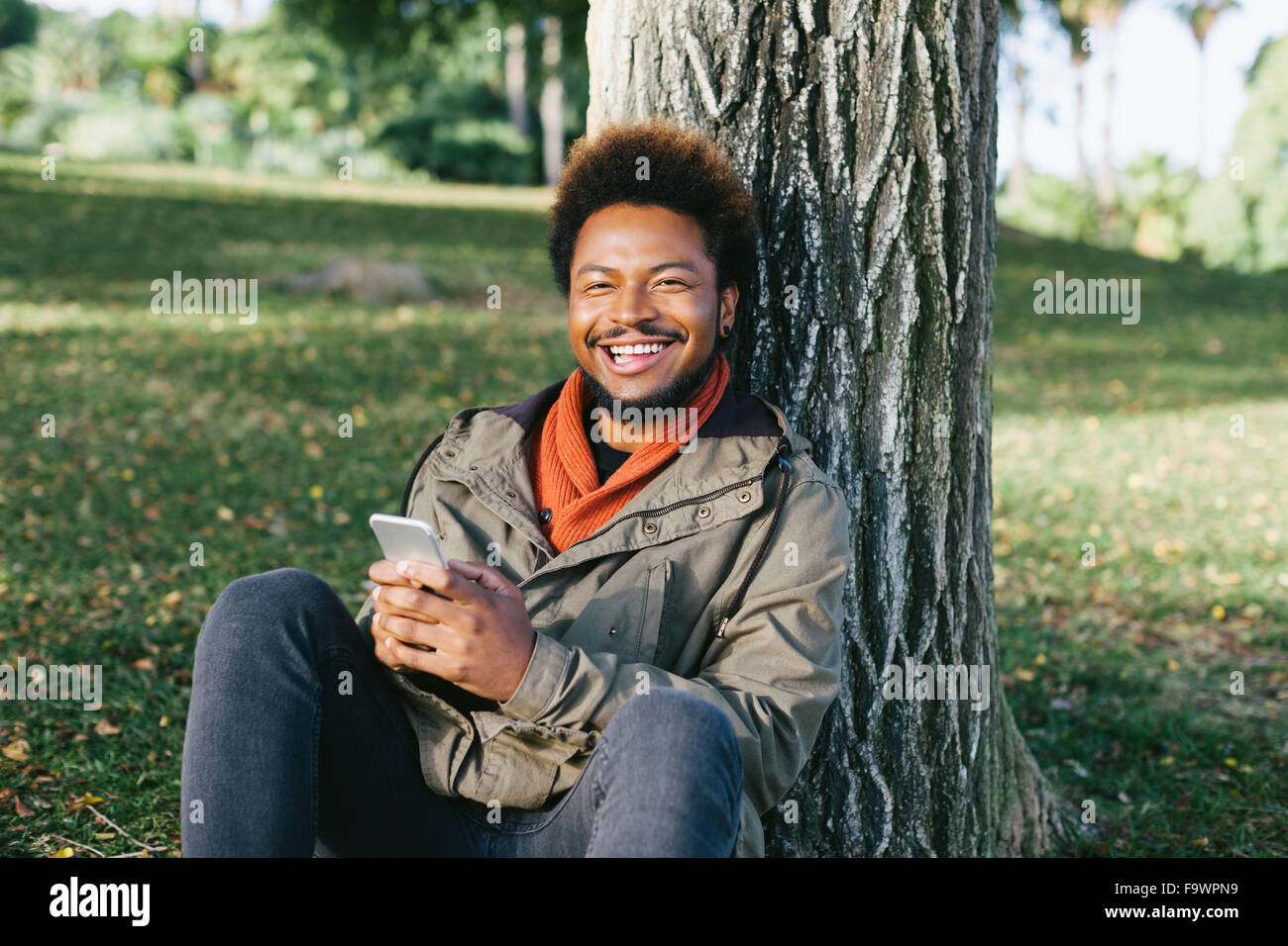 Portrait of smiling young man with smartphone leaning against tree ...