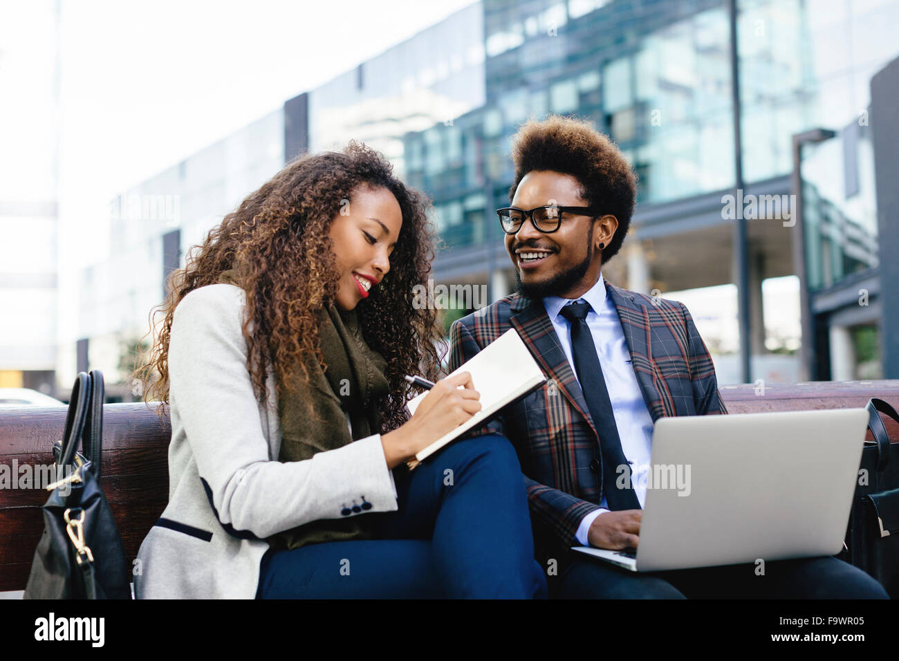 Two smiling young business people with laptop and notebook on bench ...