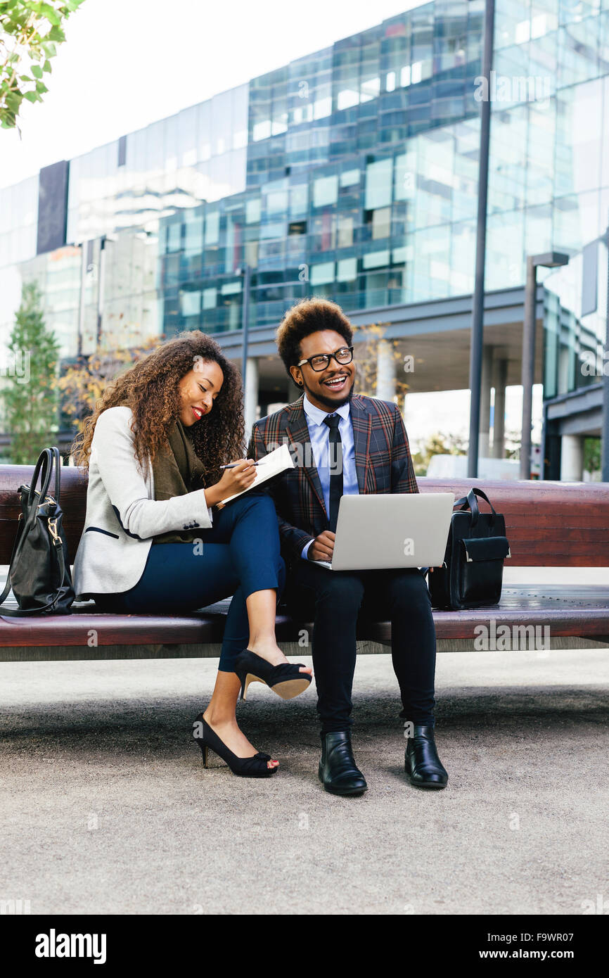 Two smiling young business people with laptop and notebook on bench ...