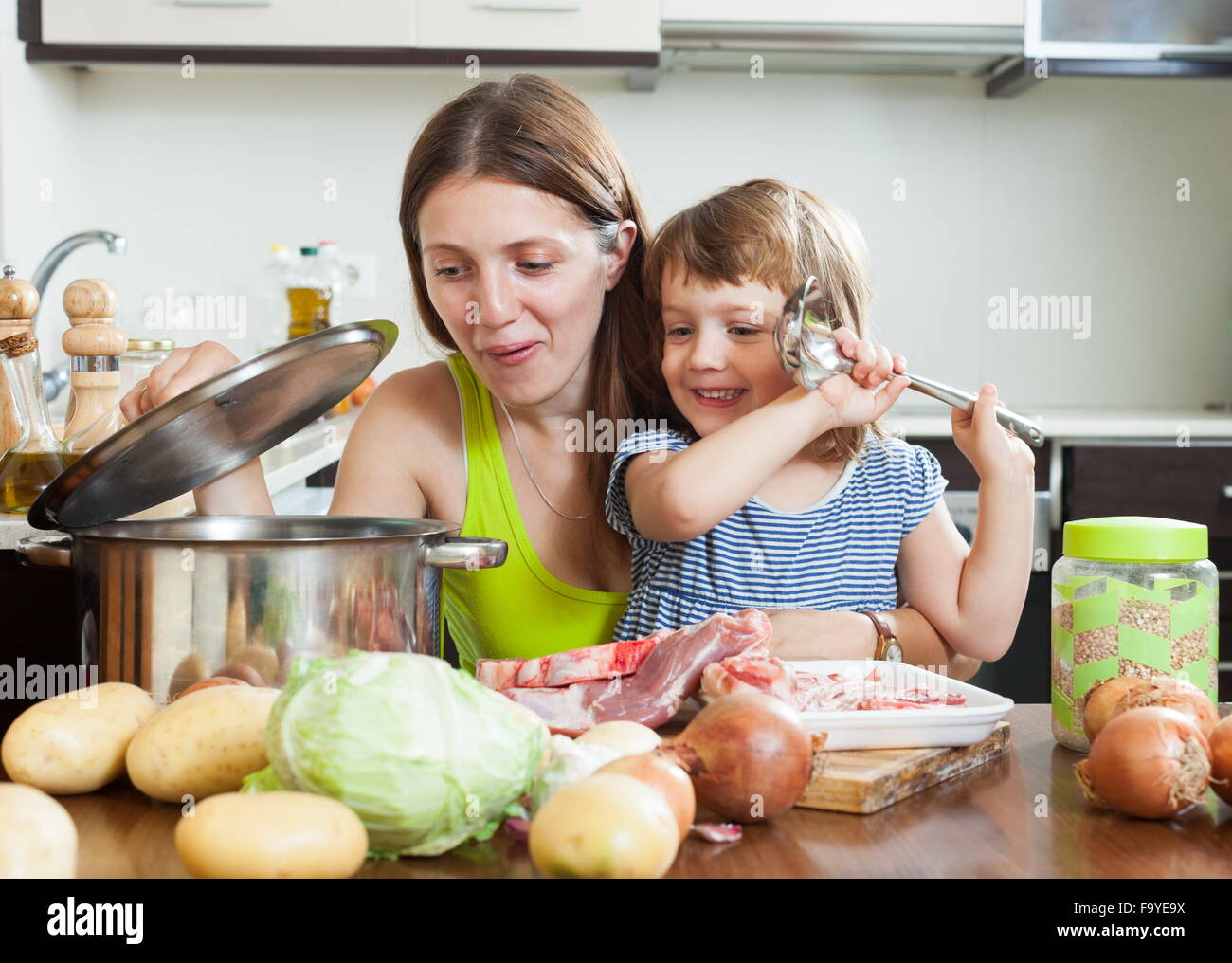 Young mother with child making soup from meat and vegetables at kitchen ...