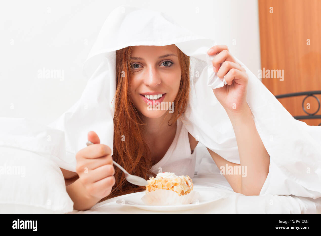 Happy woman eating sweet cake under white sheet in bed at home Stock ...