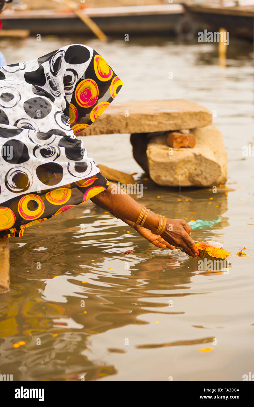 Woman at water's edge sending floating offering of flowers and sweets ...