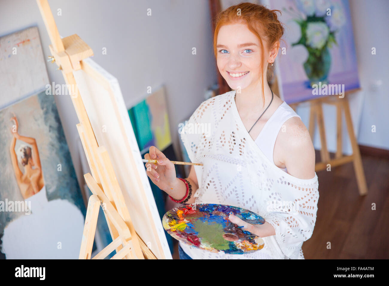 Cheerful pretty young woman artist standing and painting picture in ...