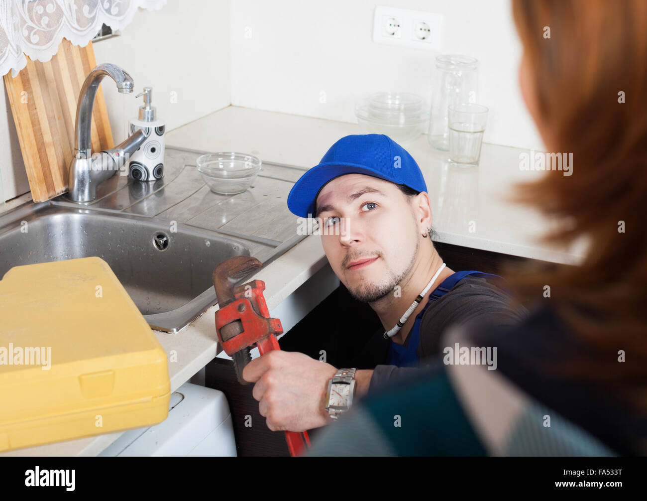 Repairman in uniform repairing a running water for housewife in kitchen ...