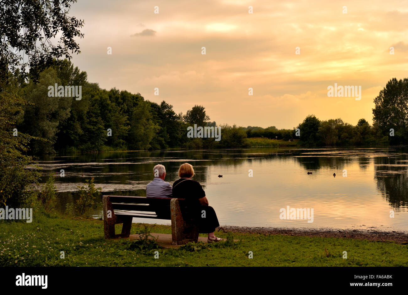 Mature couple sitting on bench overlooking lake with warm glow of ...