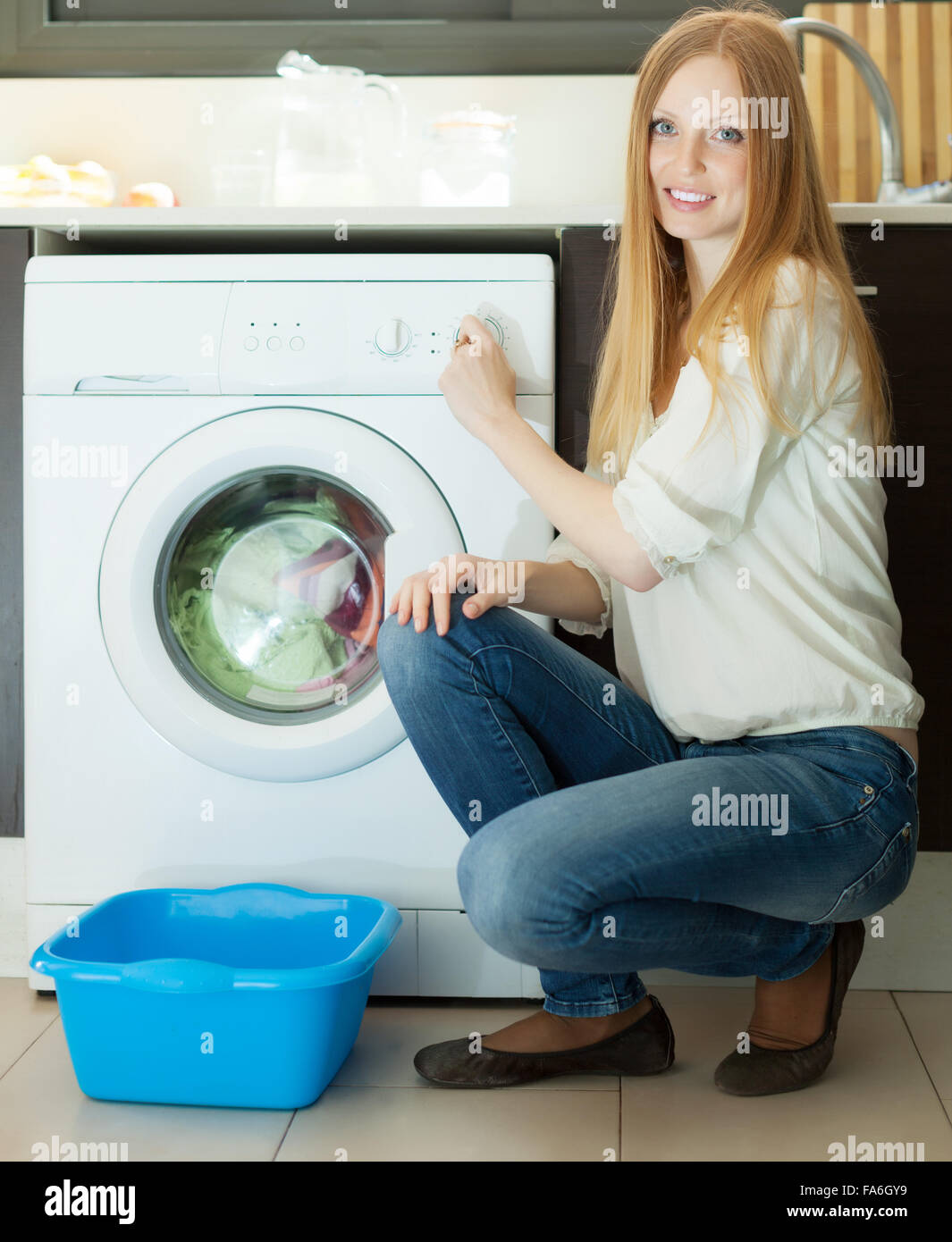 Happy long-haired woman loading clothes into the washing machine in ...