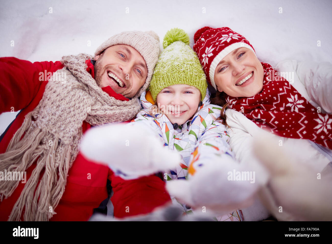 Family of three lying on snow and stretching their hands to the camera ...