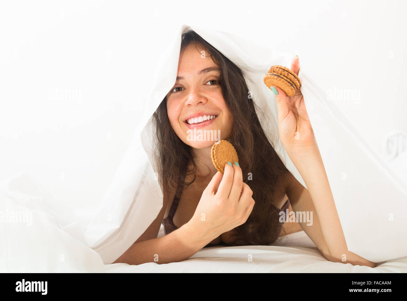 Smiling young woman eating chocolate chip cookies in her bed and ...