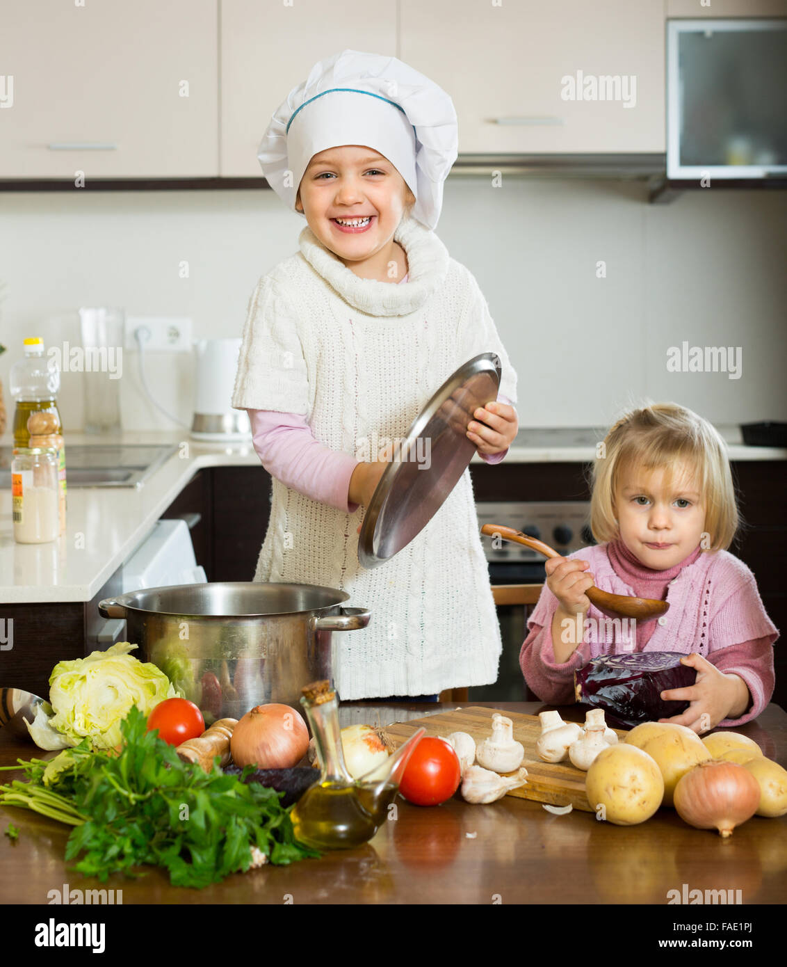 Two smiling little sisters learning how to cook at domestic kitchen ...