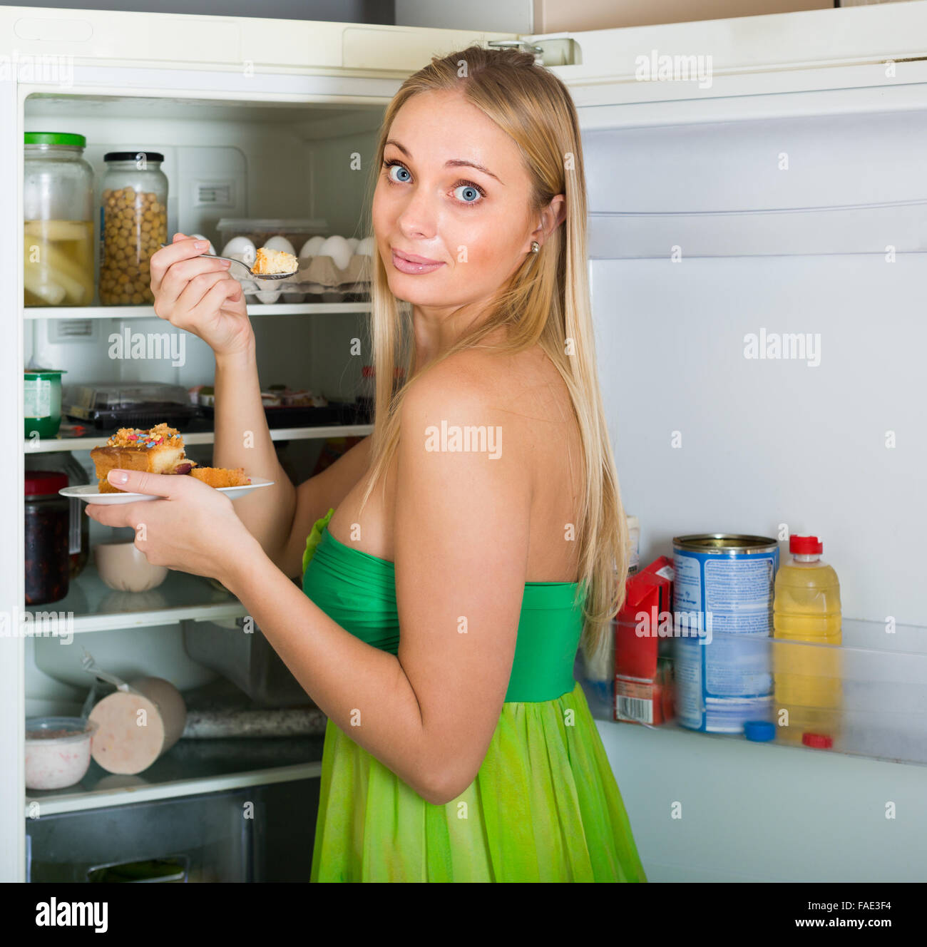 Blonde smiling girl eating biscuit from refrigerator at home Stock ...