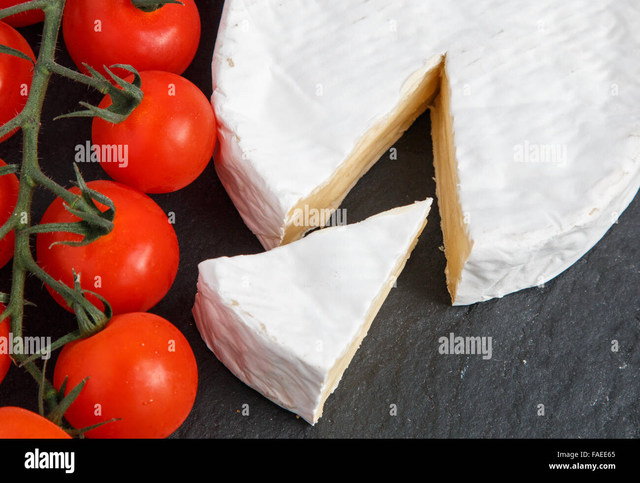 Slice of brie cut from a whole cheese next to red cherry tomatoes on a ...