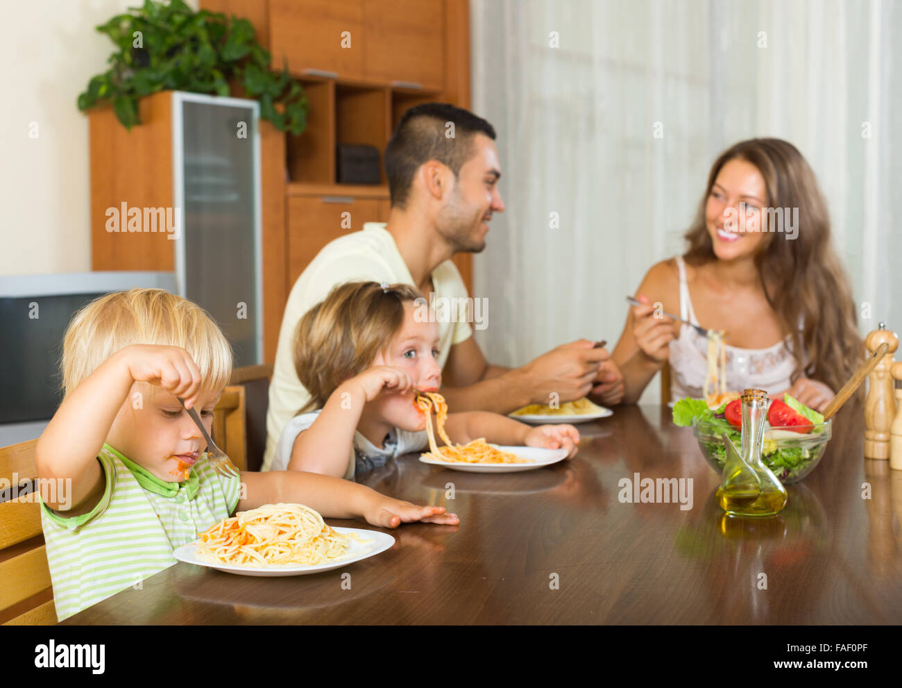 Happy young family of four eating spaghetti at home interior. Focus on ...
