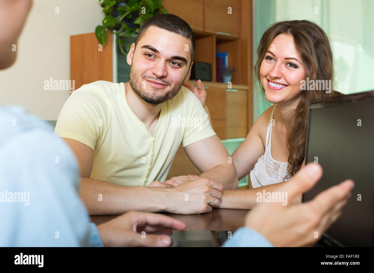 Smiling young married couple talking with employee with laptop at home ...