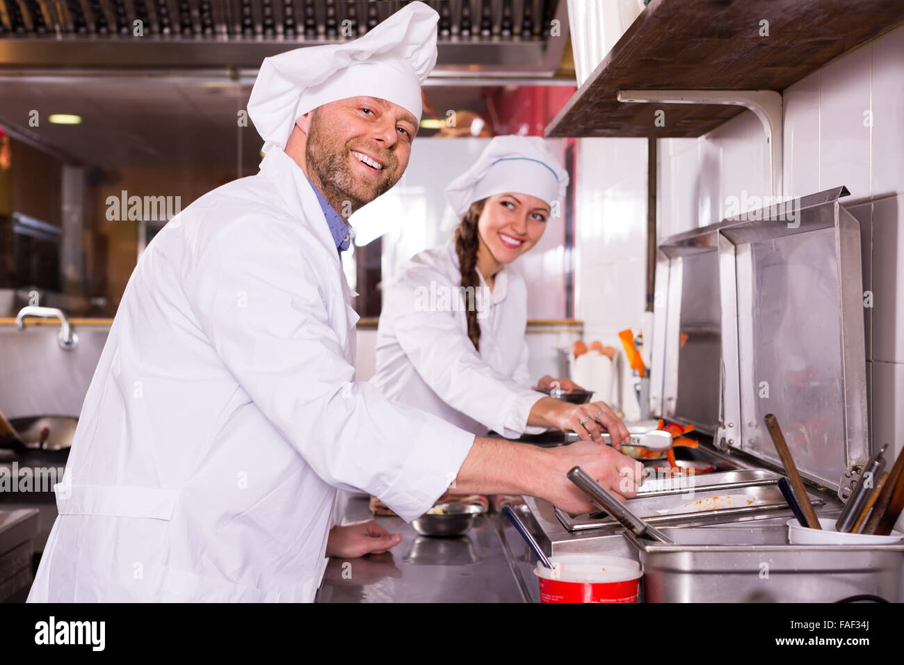 Two positive smiling cooks working together at kitchen in take-away ...