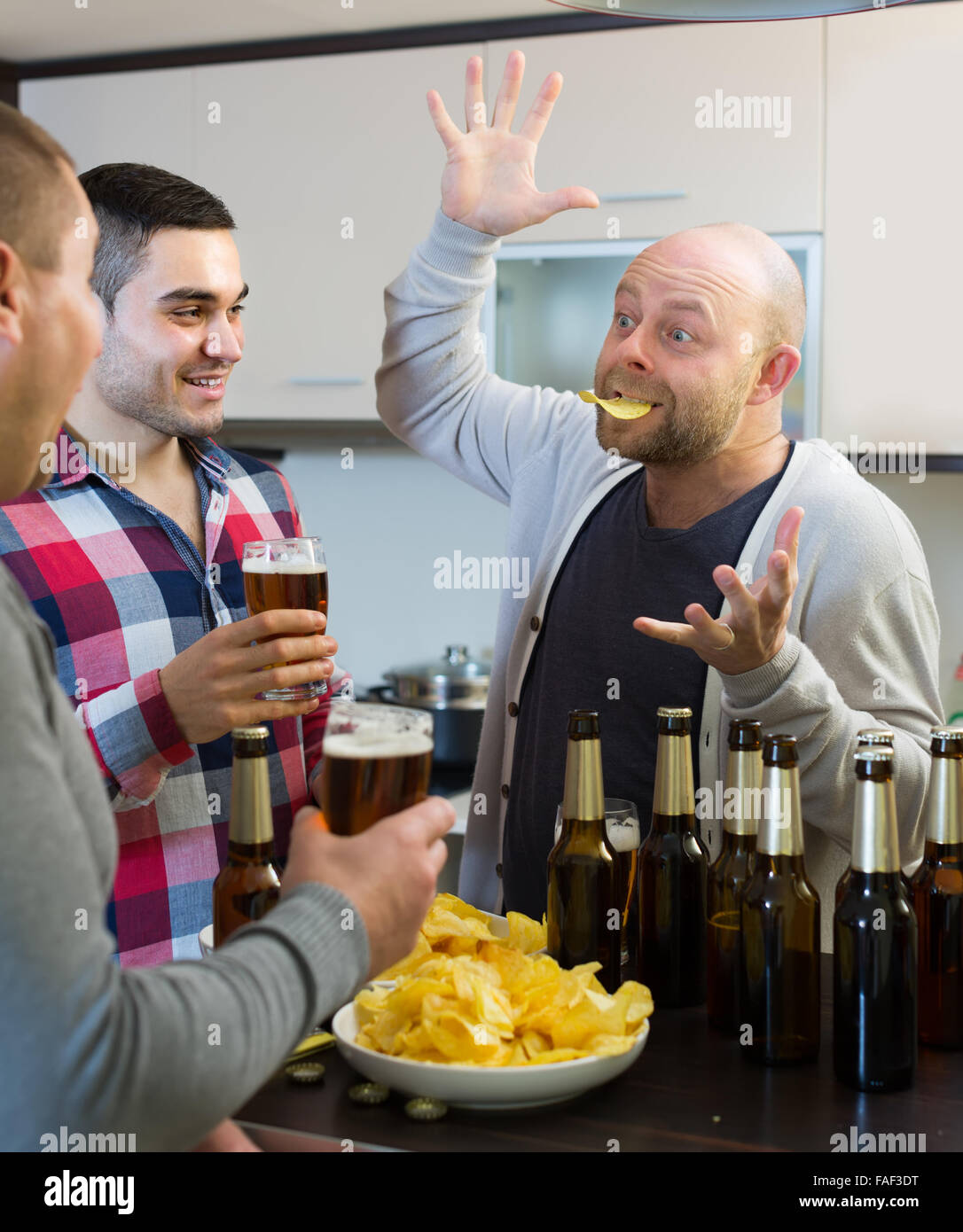 Three smiling male friends chatting at the kitchen with glasses of beer ...