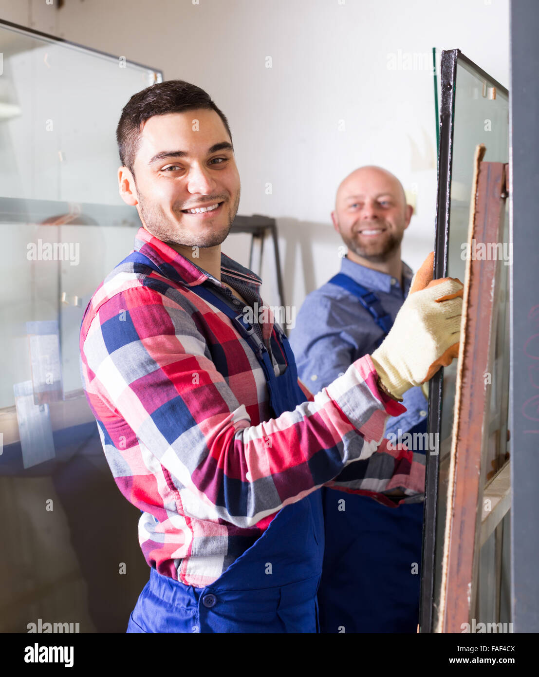Two positive smiling workmen in coverall working with glass at factory ...