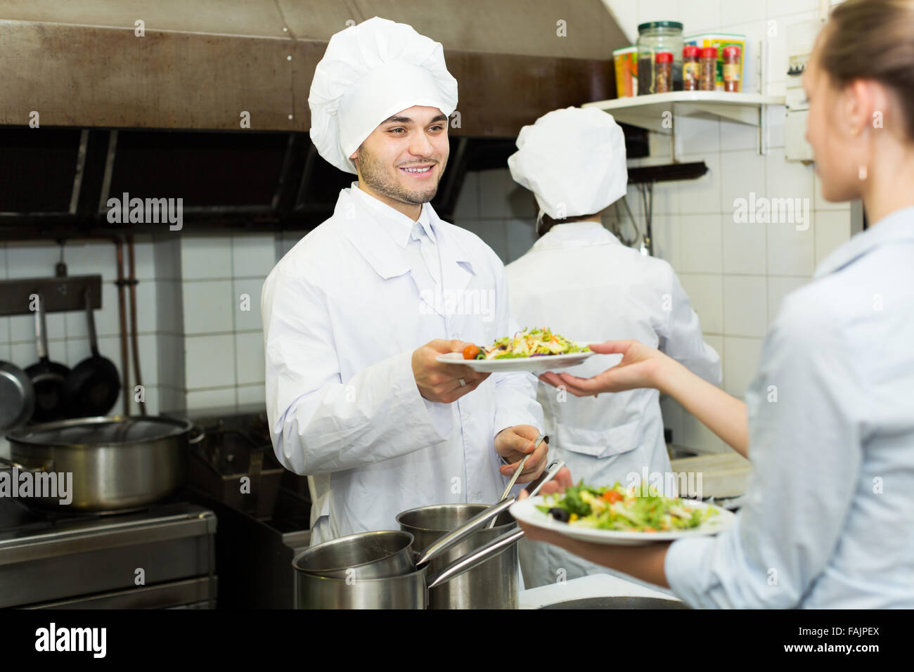 Positive young cook gives to female waitress plates with prepared meal ...