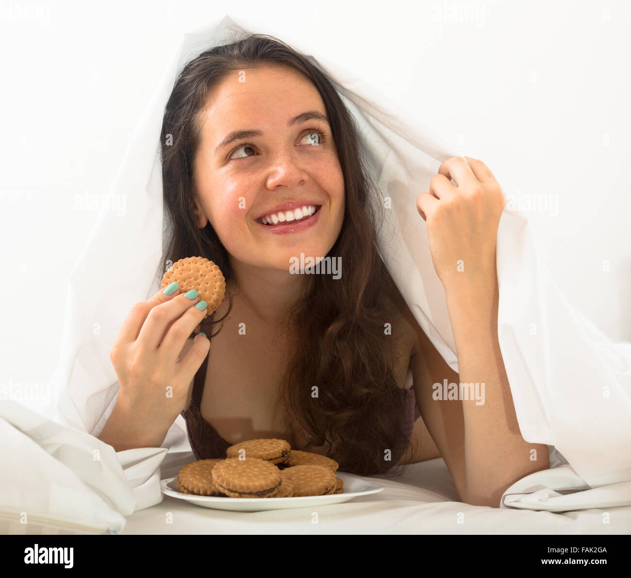 Smiling adult girl eating chocolate chip cookies in her bed at home ...