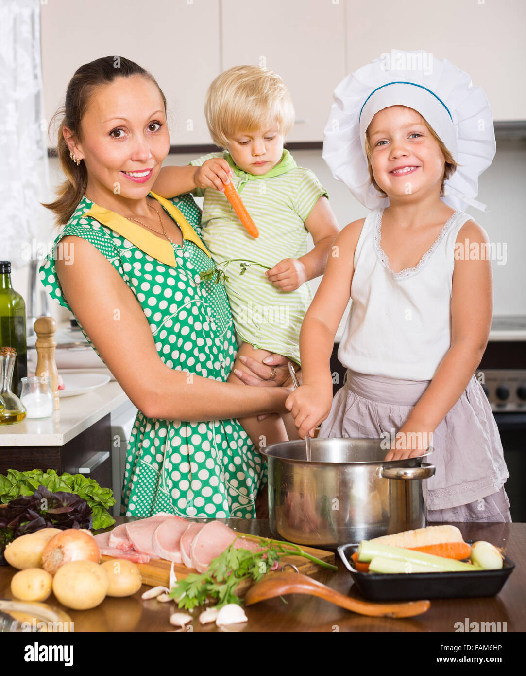 Smiling mother with baby in arms and little daughter cooking together ...