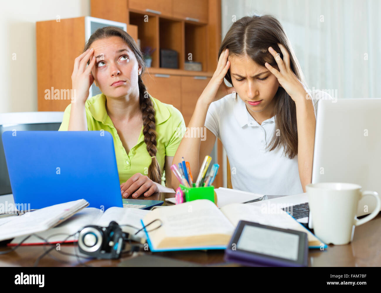 Portrait of two tired girls sitting at the table with laptops and books ...