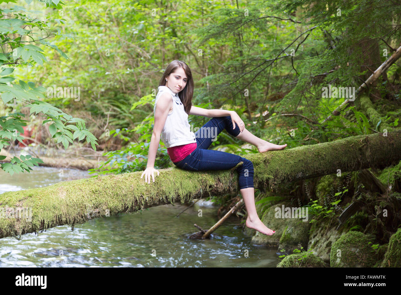 Portrait of a teenage girl sitting on a moss covered log over a stream ...