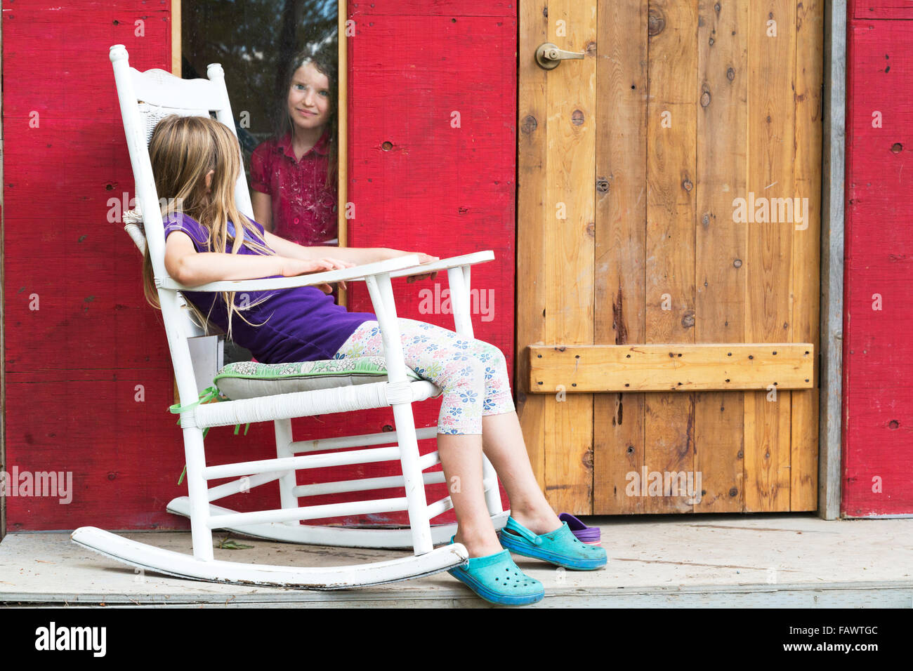 A young girl looks out a window at another girl sitting in a rocking ...