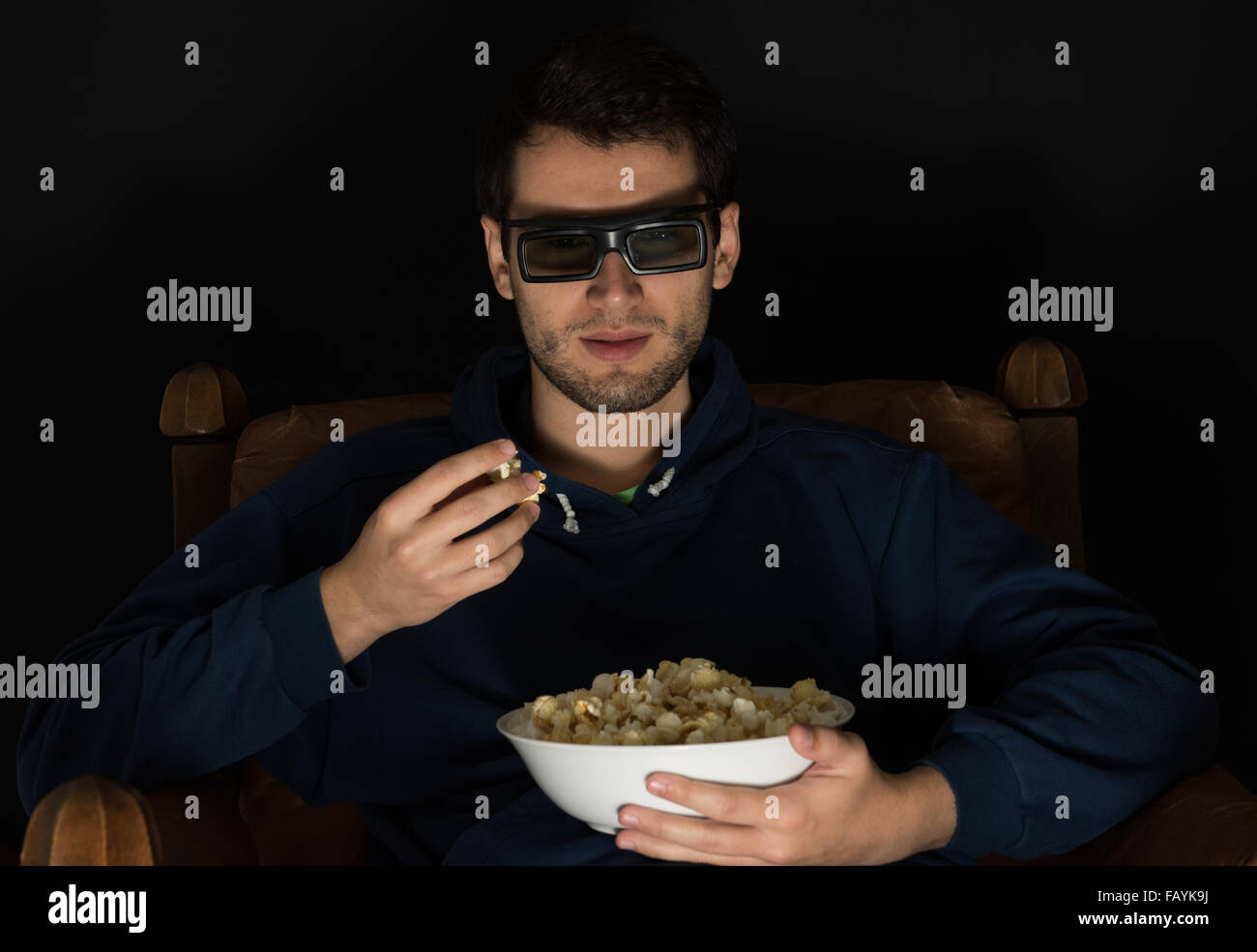 young man sitting in the dark room in the front of tv watching movie and eating popcorn, wearing ...