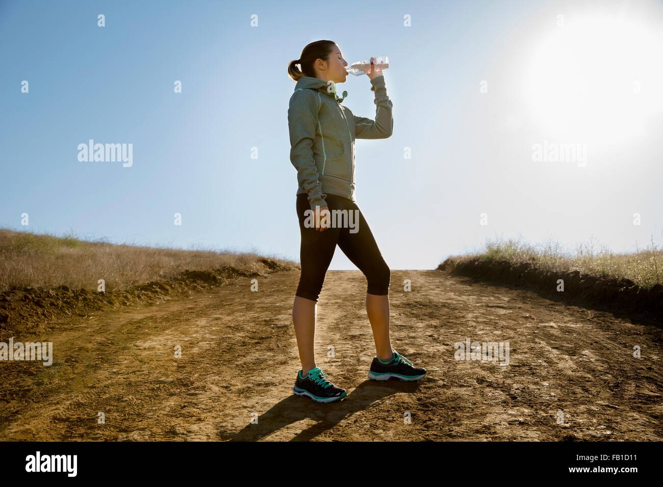 Silhouetted female runner drinking from water bottle on dirt track ...
