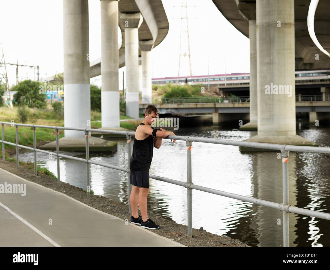 Young man beside river, leaning on railings, using MP3 player attached ...