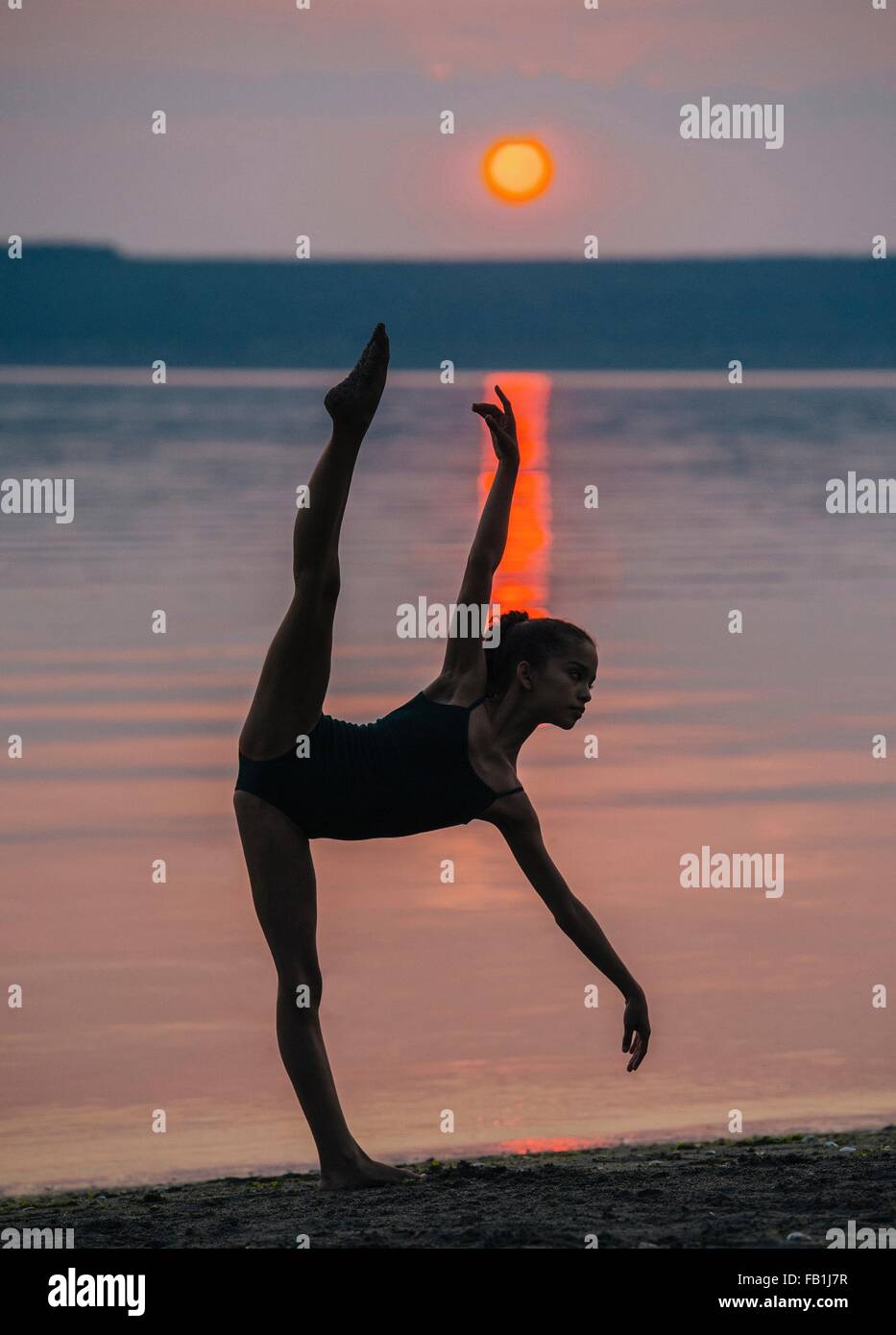 Girl by ocean at sunset on one leg, bending forward arm and leg raised ...