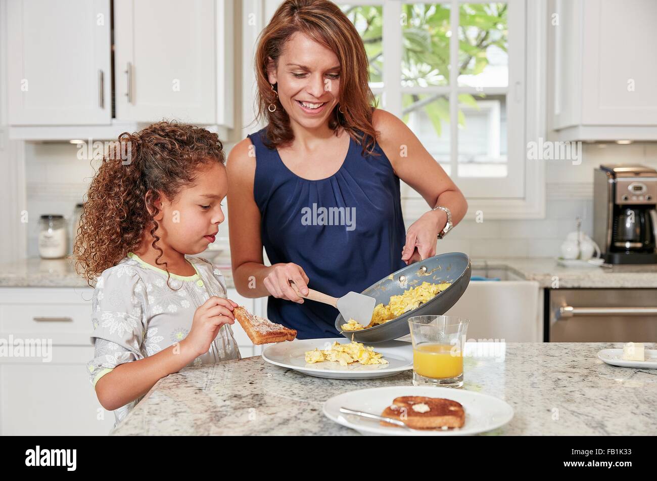 Mother at kitchen counter serving daughter scrambled eggs from frying ...