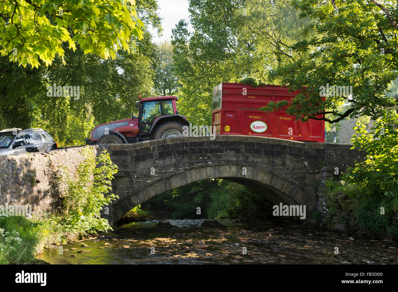 Tractor pulling a red trailer drives over an old, rustic, stone bridge ...