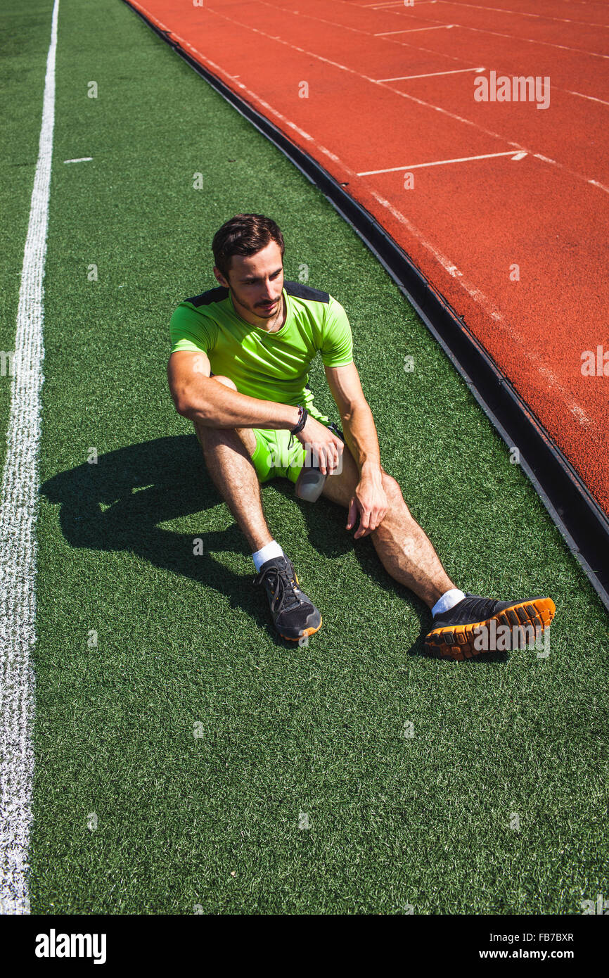 Full length of male athlete with water bottle relaxing on sports field ...