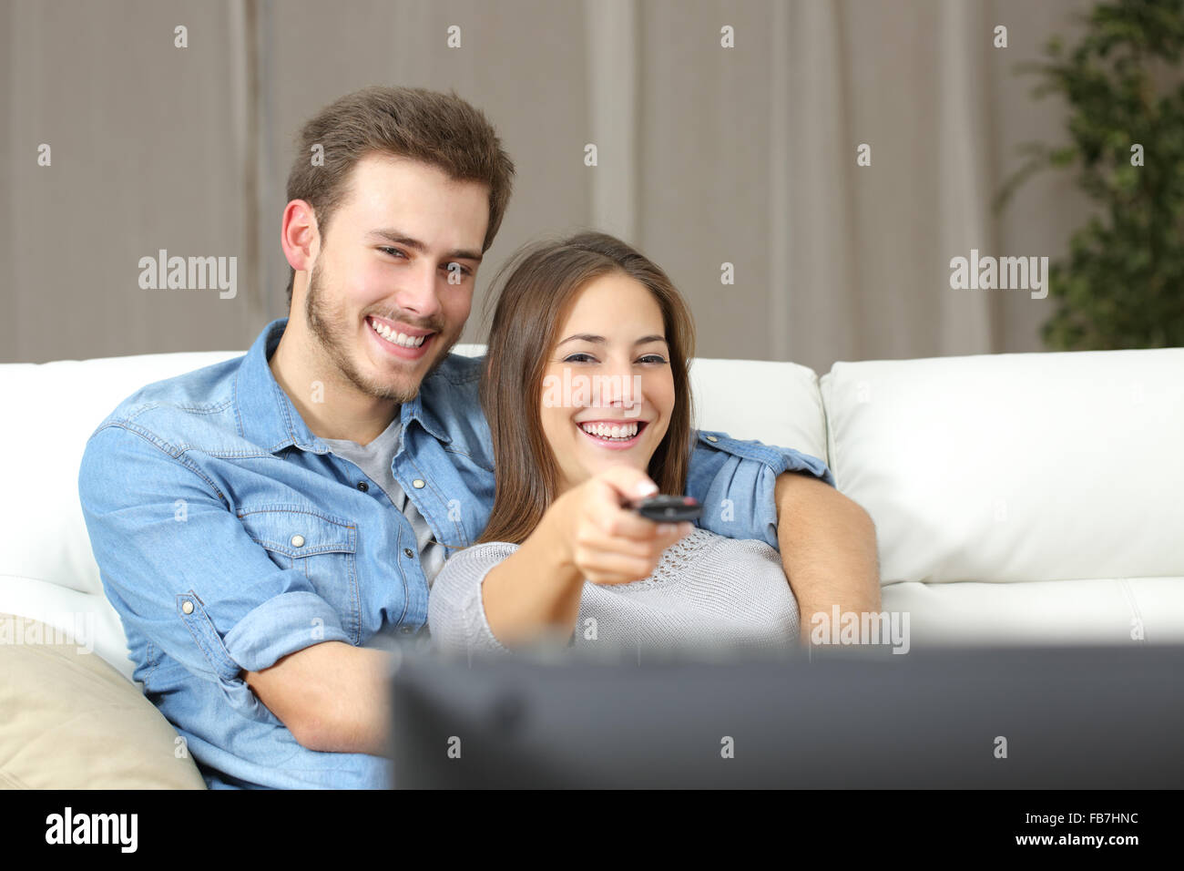 Happy couple using remote control to change channel on tv sitting on a ...