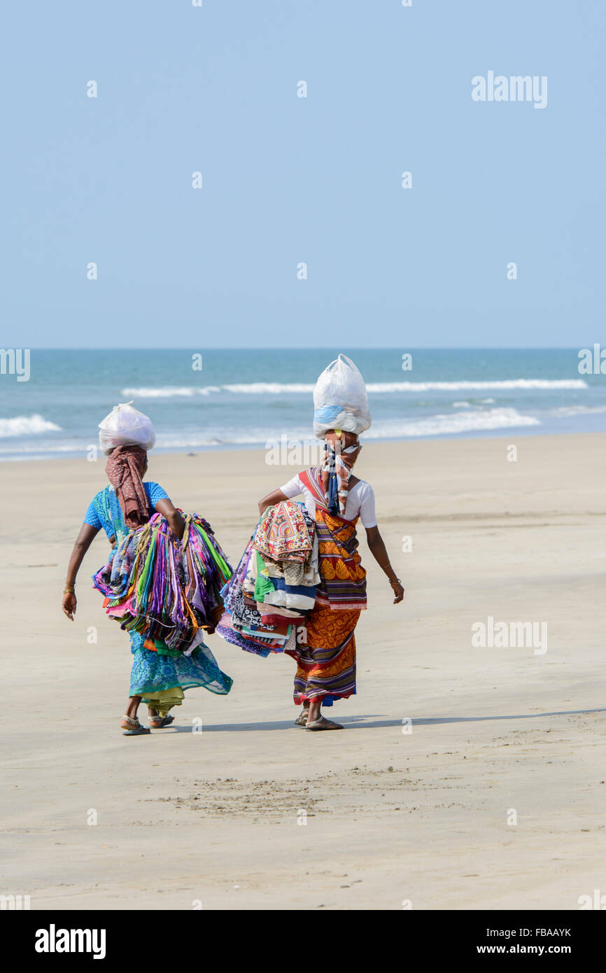 Two Indian women selling sarongs and fabrics walk along an empty beach ...