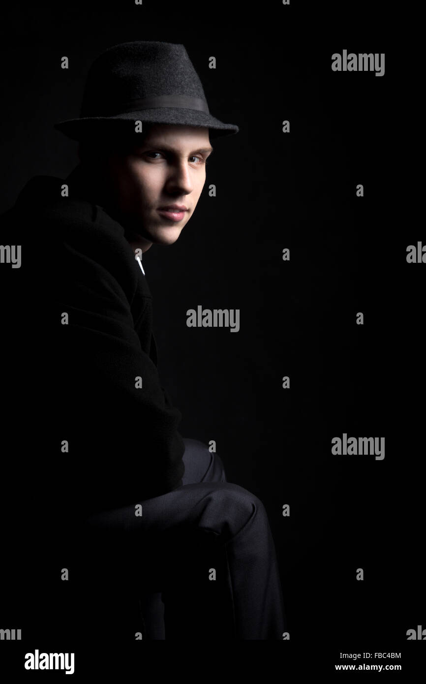 Low key shot of mysterious young man wearing hat and coat sitting in ...