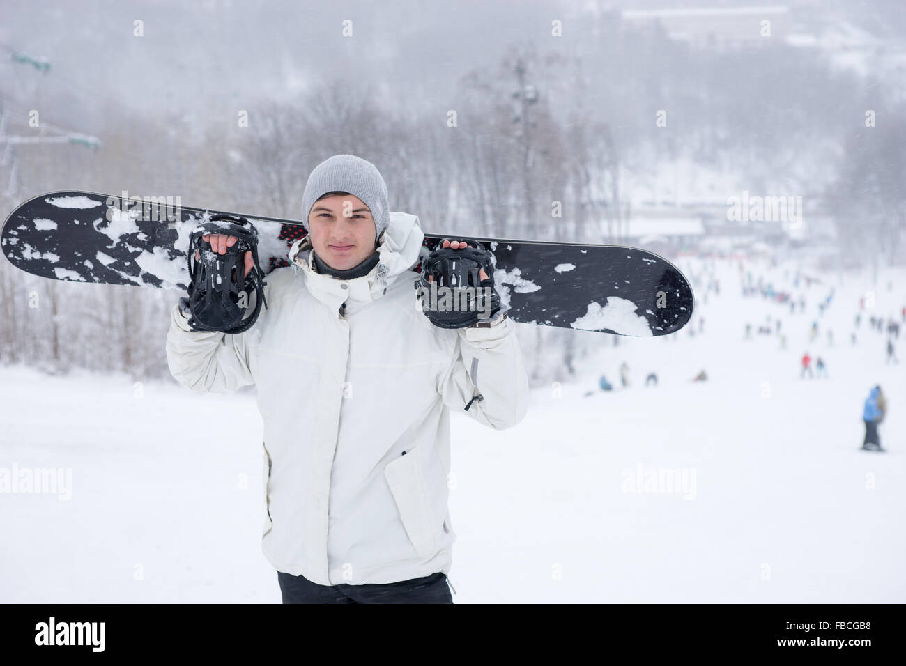Smiling friendly young male snowboarder standing with his board across ...