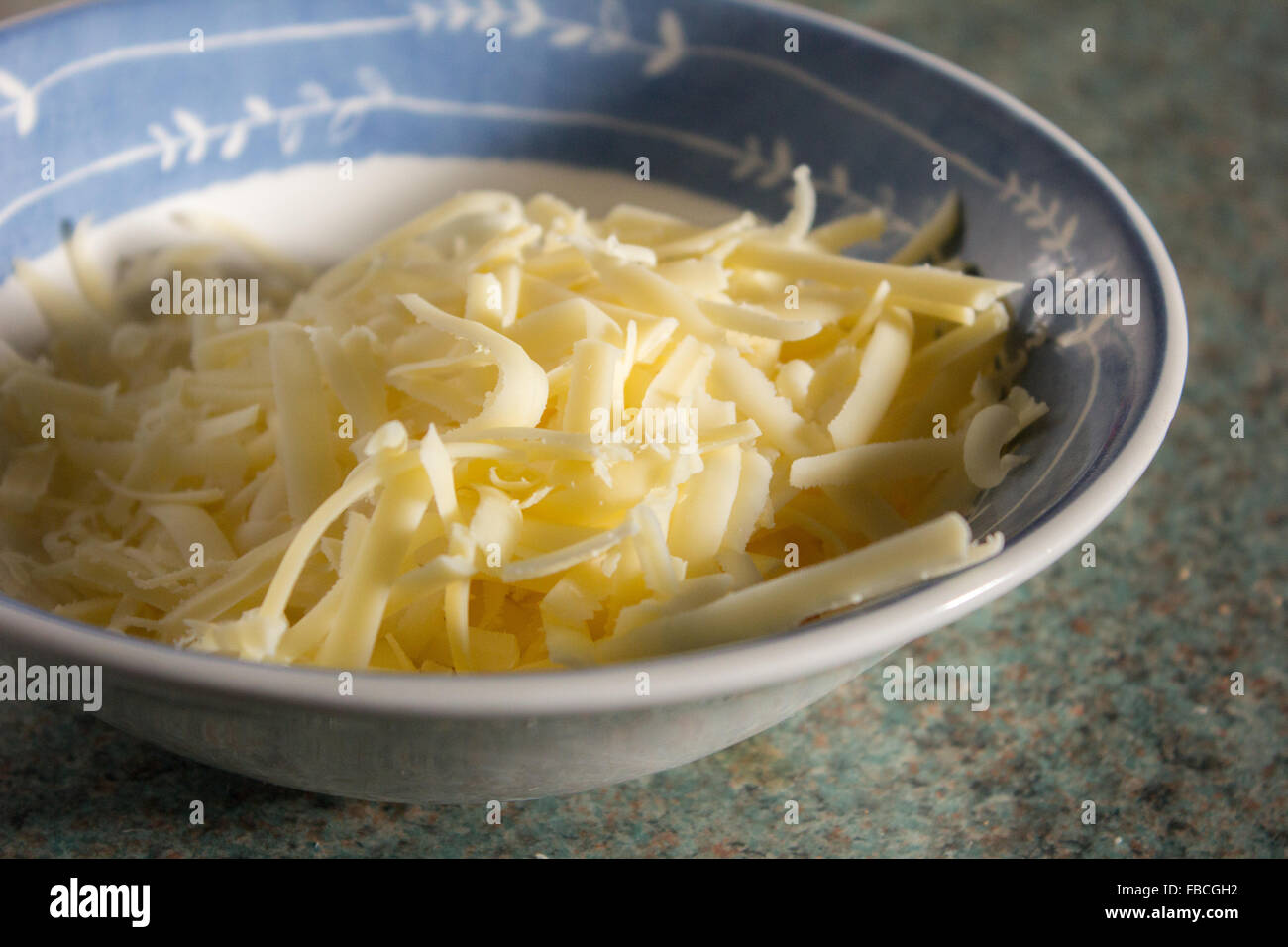 Grated cheddar cheese in a natural lit bowl on a mottled green kitchen ...